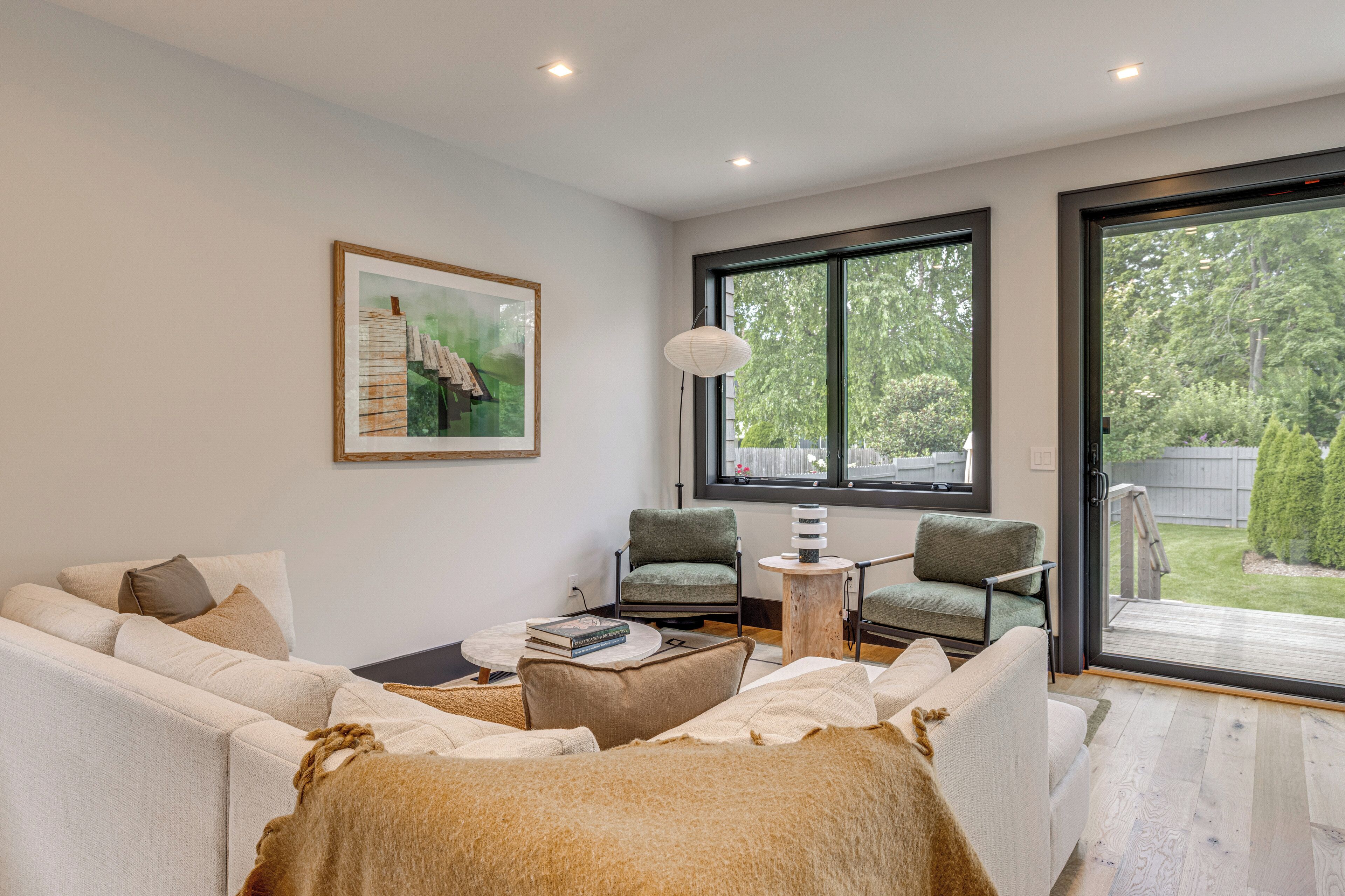 Uma sala de estar moderna com um conjunto de sofá, uma mesa de centro e duas cadeiras. Há um quadro na parede e uma janela com vista para o verde lá fora.