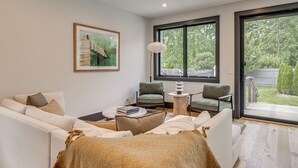 Uma sala de estar moderna com um conjunto de sofá, uma mesa de centro e duas cadeiras. Há um quadro na parede e uma janela com vista para o verde lá fora.