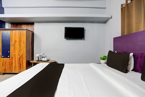 Quarto de hotel com cama, televisão na parede, guarda-roupa de madeira, mesa pequena com chaleira e chávenas, e planta em vaso.
