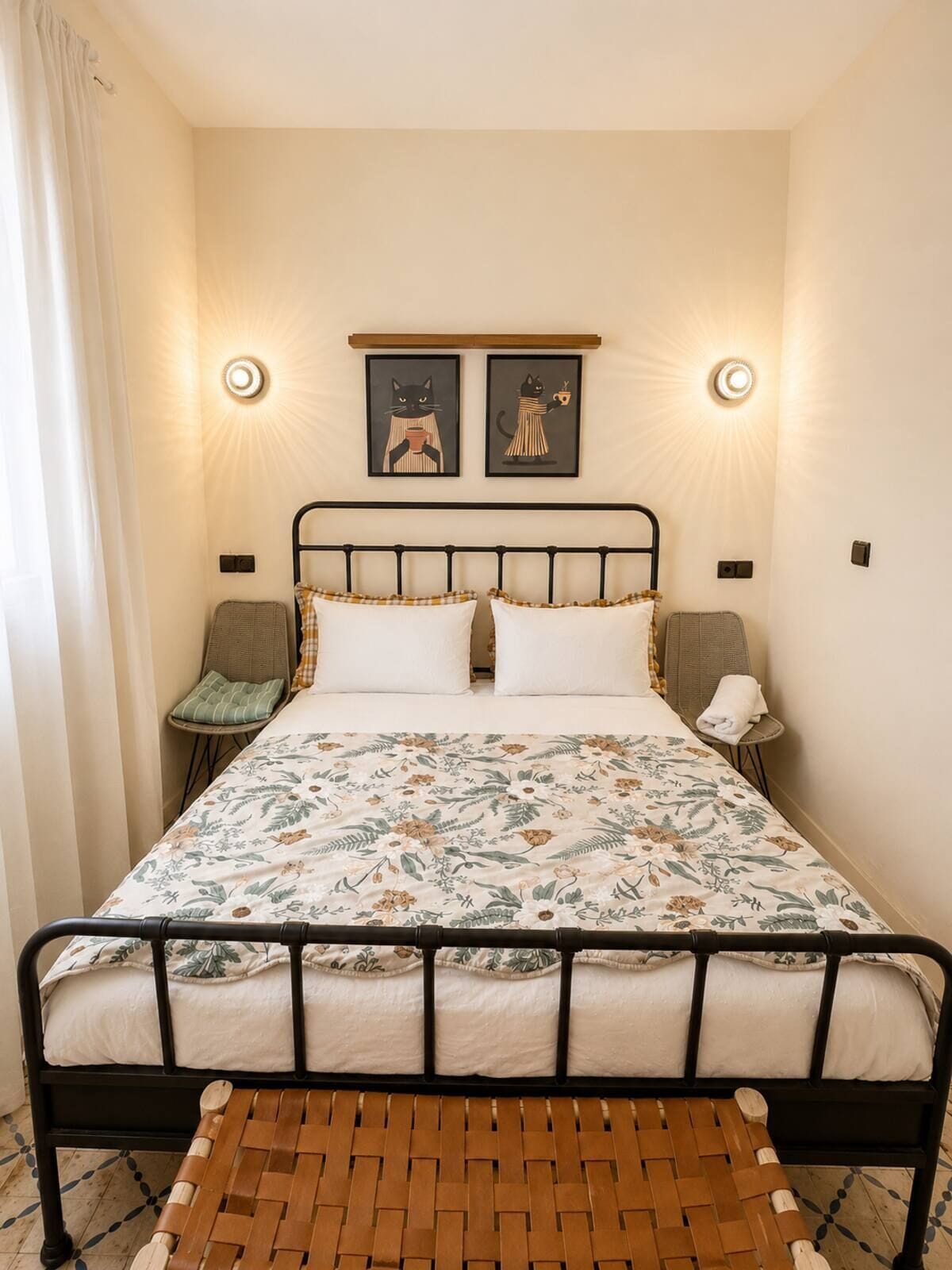 Uma cama com colcha florida, duas luminárias fixadas na parede e duas fotos emolduradas sobre a cama.