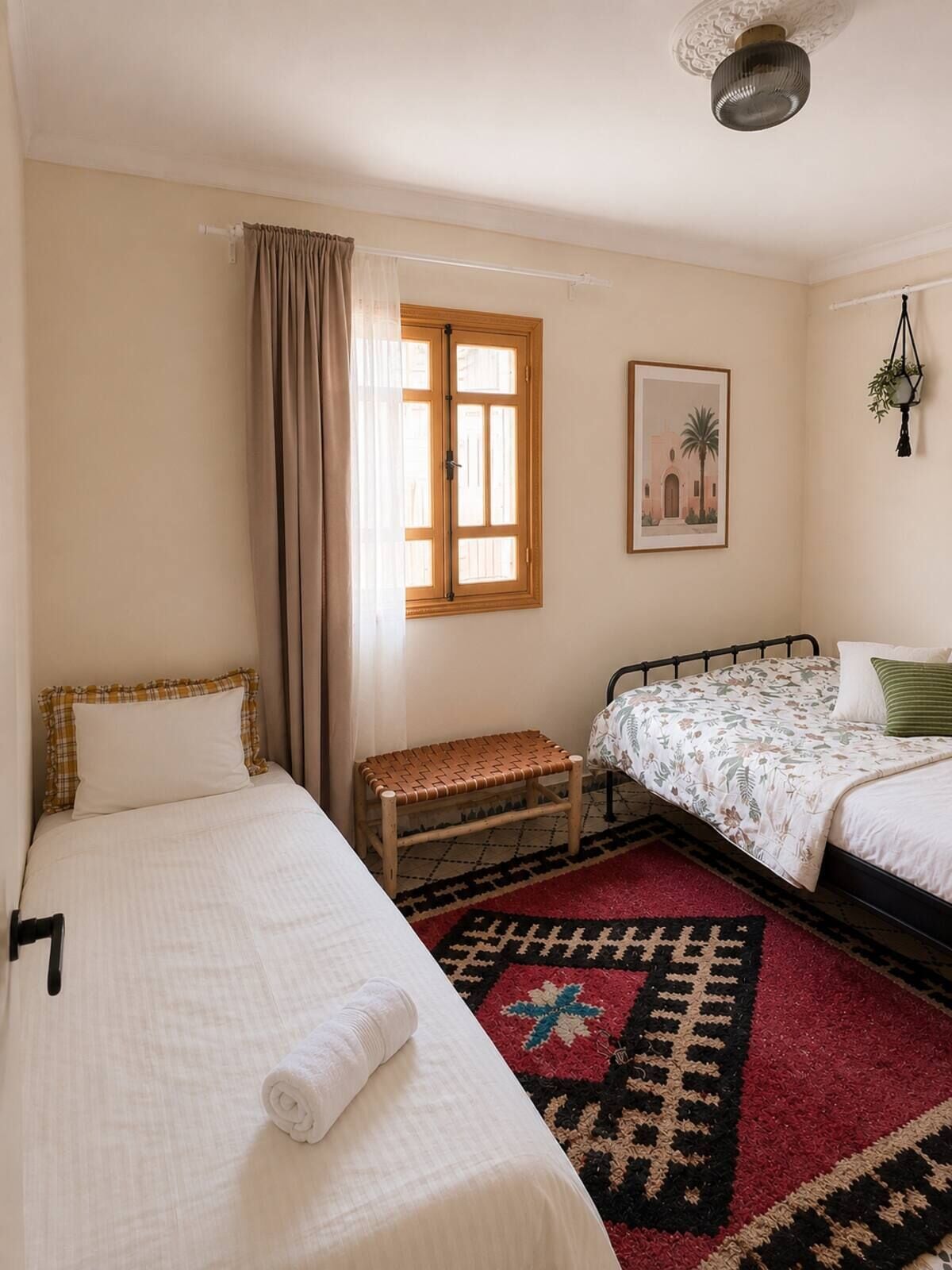 Quarto com duas camas, um banquinho, janela com cortinas, planta pendente e tapete estampado.