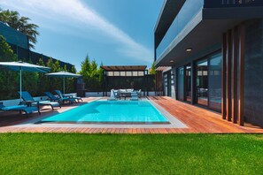 Uma área de piscina moderna ao ar livre com espreguiçadeiras e guarda-sóis, cercada por um deck de madeira e um gramado.