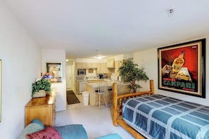 Quarto pequeno com cama, escrivaninha, cadeira e cozinha compacta.