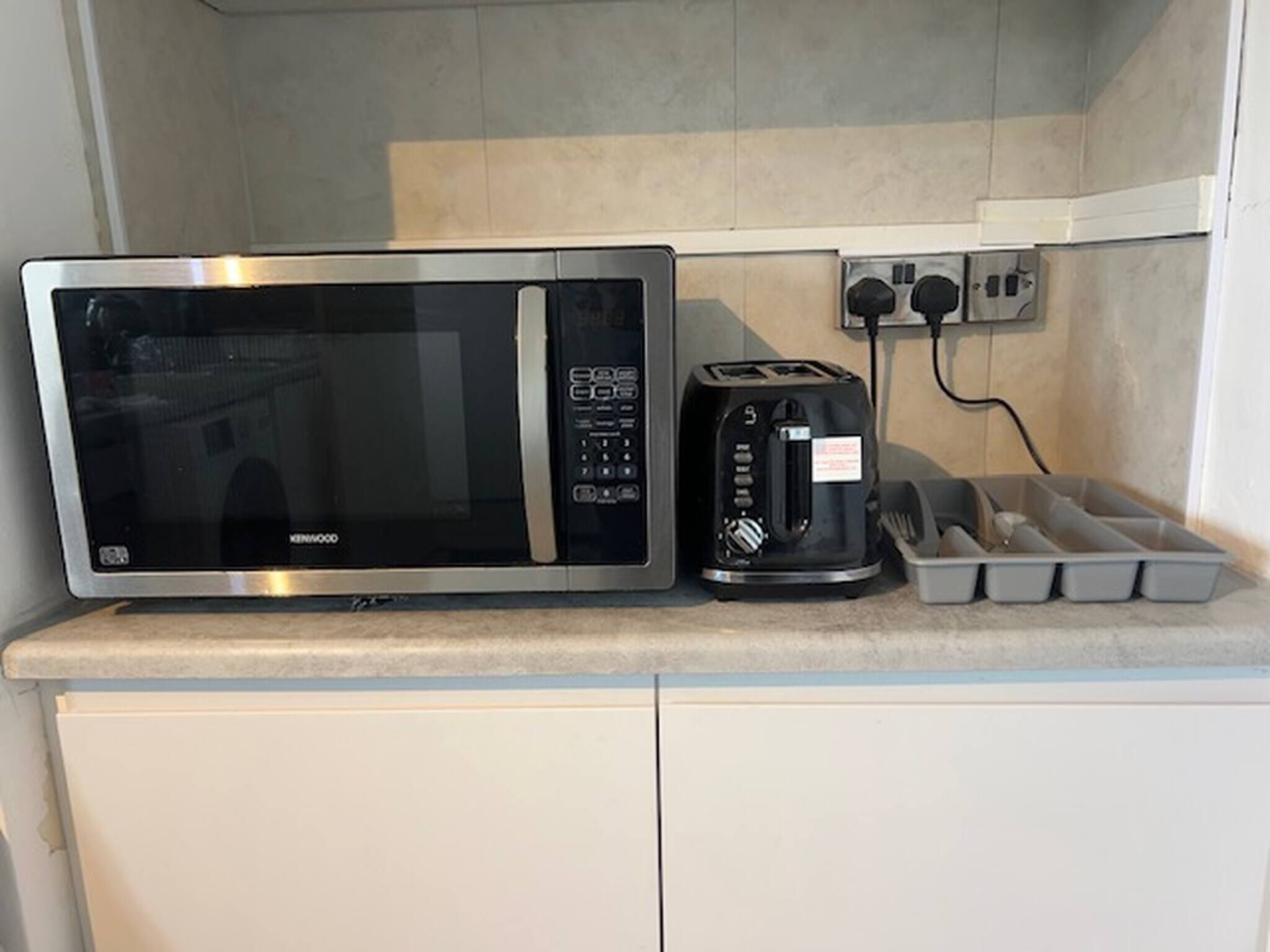 Um forno micro-ondas com painel de controle digital, uma cafeteira e um conjunto de quatro bandejas cinzas em uma bancada.