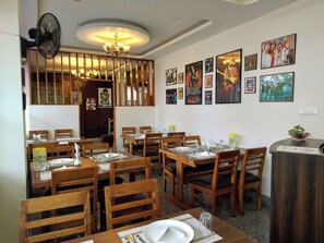 Interior de restaurante com mesas e cadeiras de madeira, um ventilador e obras de arte nas paredes.