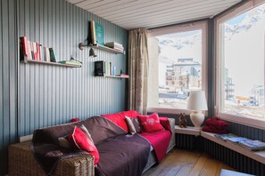 Um quarto aconchegante com piso de madeira, um sofá vermelho com almofadas, uma estante de livros, uma janela com vista para edifícios e um abajur sobre uma mesa lateral.