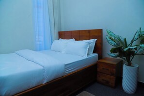 Uma cama bem arrumada com roupa de cama branca, cabeceira de madeira, mesa de cabeceira com gavetas e um vaso com planta de folhas variegadas.