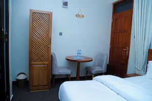 Quarto de hotel com cama, duas cadeiras, uma mesinha com garrafas de água, um armário de madeira e uma porta.