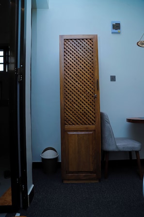 Um quarto com porta de madeira, uma cadeira cinza, uma mesinha e uma lixeira.