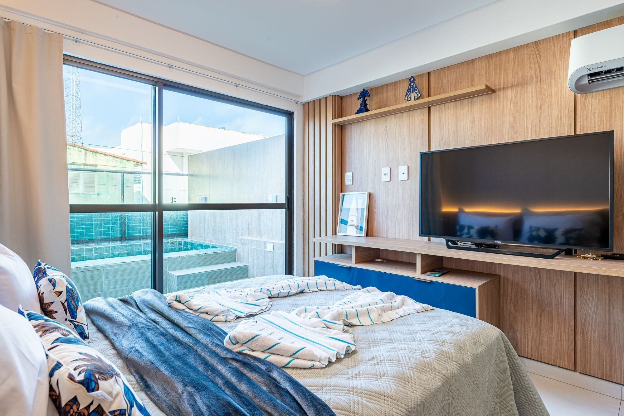 Quarto de hotel com uma cama grande, televisão e varanda com vista para edifícios.