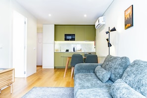 Uma sala de estar moderna com um sofá cinzento e um banco de madeira, uma cozinha com armários verdes e um micro-ondas, e um pôster emoldurado na parede.