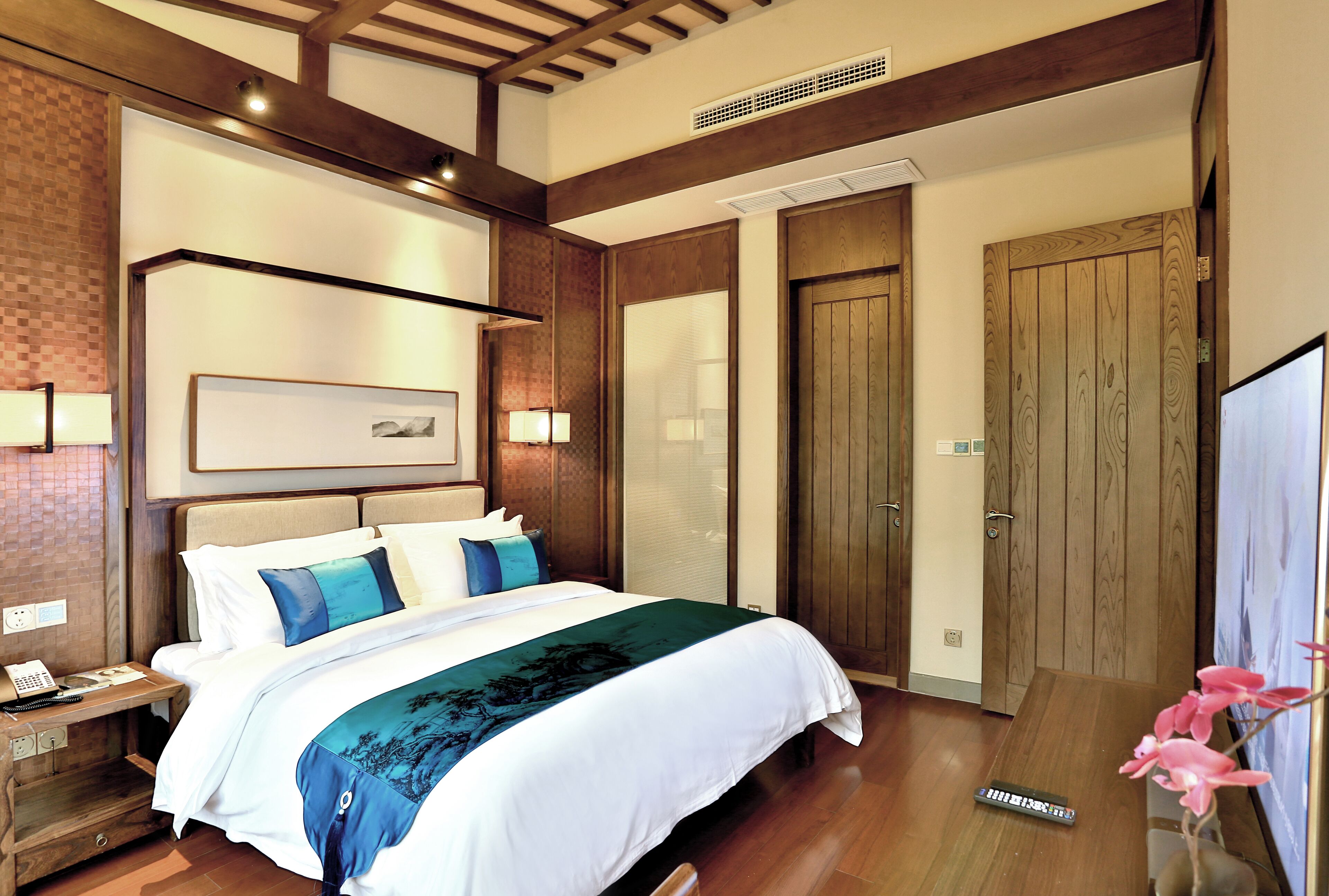 Quarto de hotel com uma cama grande, mesa de cabeceira, televisão na parede e porta de madeira.