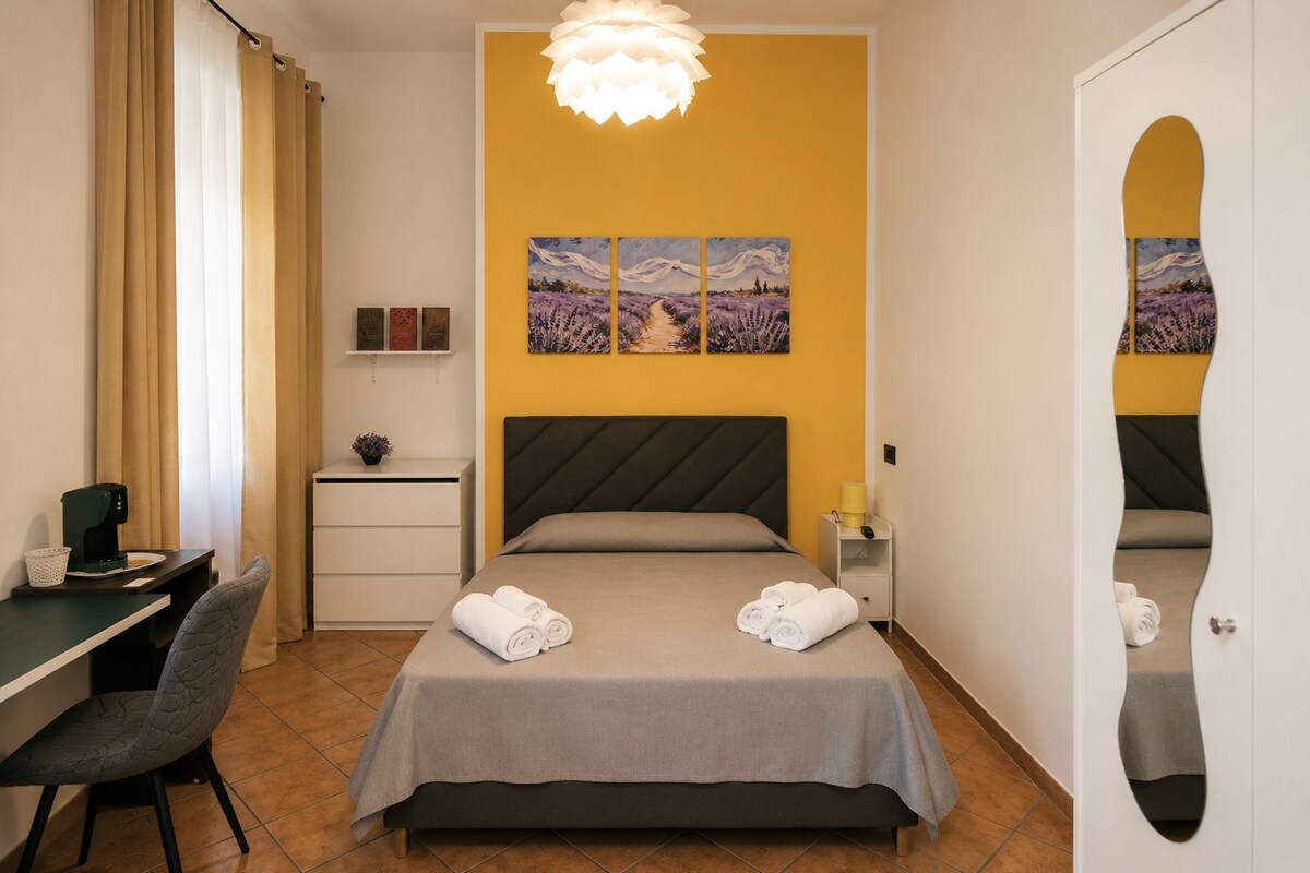 Quarto de hotel com cama, escrivaninha, cadeira, mesa de cabeceira, abajur e obras de arte na parede.