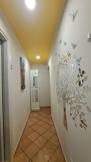 Um corredor com piso de azulejos, uma porta com a inscrição '2', uma parede com um mural de árvore e borboleta, e uma porta com espelho e decoração de borboleta.