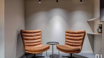 Quarto moderno com duas poltronas de couro e uma mesa redonda pequena, uma máquina de café em uma prateleira, e teto visível com luminárias.