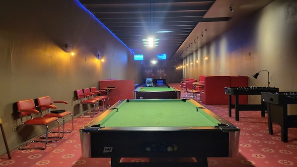 Uma sala de bilhar com mesa de sinuca, assentos vermelhos e uma pista de boliche ao fundo.