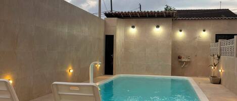 Piscina com espreguiçadeiras e uma mesa, cercada por um muro baixo e um edifício com telhado telhado.