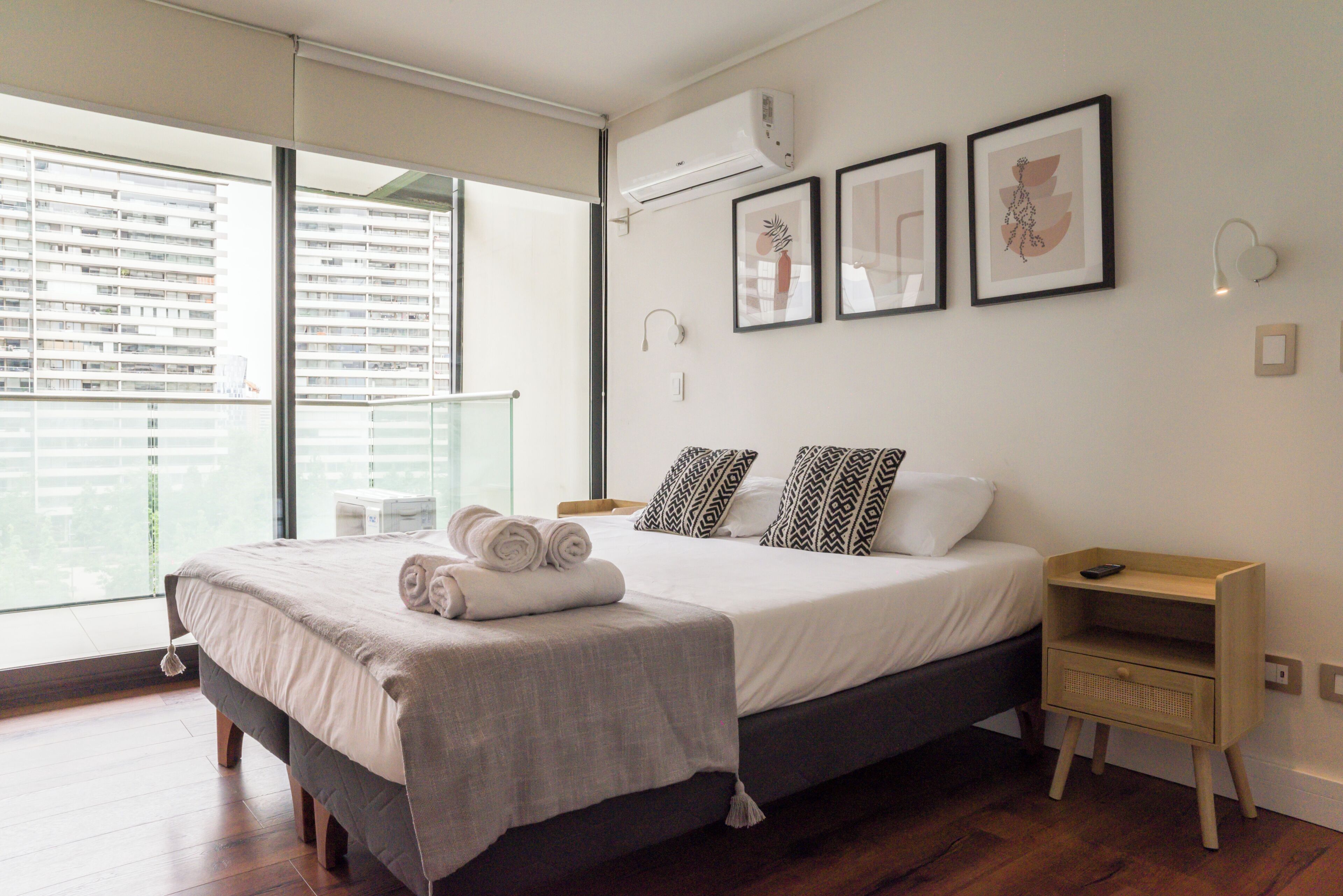Quarto de hotel moderno com uma cama grande, uma mesa de cabeceira, um criado-mudo de madeira e uma janela com vista para um edifício alto.