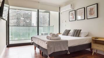Quarto moderno com cama grande, mesa de cabeceira, janela com vista para edifícios altos e três obras de arte emolduradas na parede.