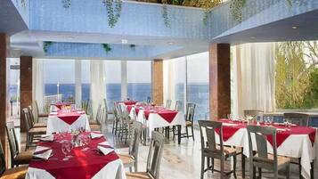 Restaurante com mesas montadas para refeições, amplas janelas com vista para o mar e teto alto com plantas pendentes.