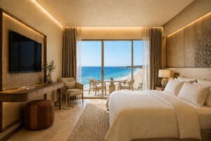 Quarto de hotel com uma cama grande, uma escrivaninha com cadeira, televisão e vista para a praia.