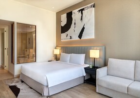 Quarto de hotel com uma cama grande, duas mesas de cabeceira com abajures, um sofá e um quadro abstrato de grandes dimensões na parede.