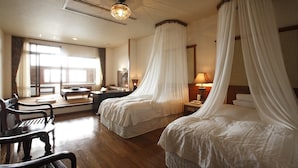 Quarto de hotel com duas camas, um espaço de estar com mesa e cadeiras, uma janela ampla e um ventilador de teto.