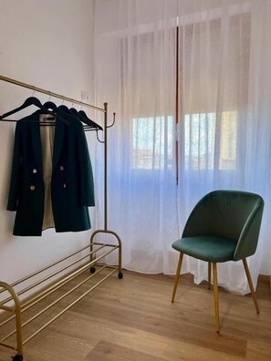 Quarto com um cabideiro, uma cadeira e uma janela com cortinas translúcidas.