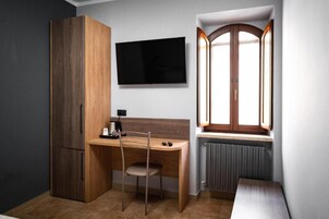 Um quarto pequeno com uma escrivaninha de madeira, uma cadeira, uma televisão na parede e uma janela com vista.