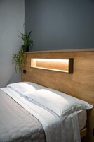 Uma cama com cabeceira de madeira, uma planta na mesinha de cabeceira e um luminário fixado na parede.