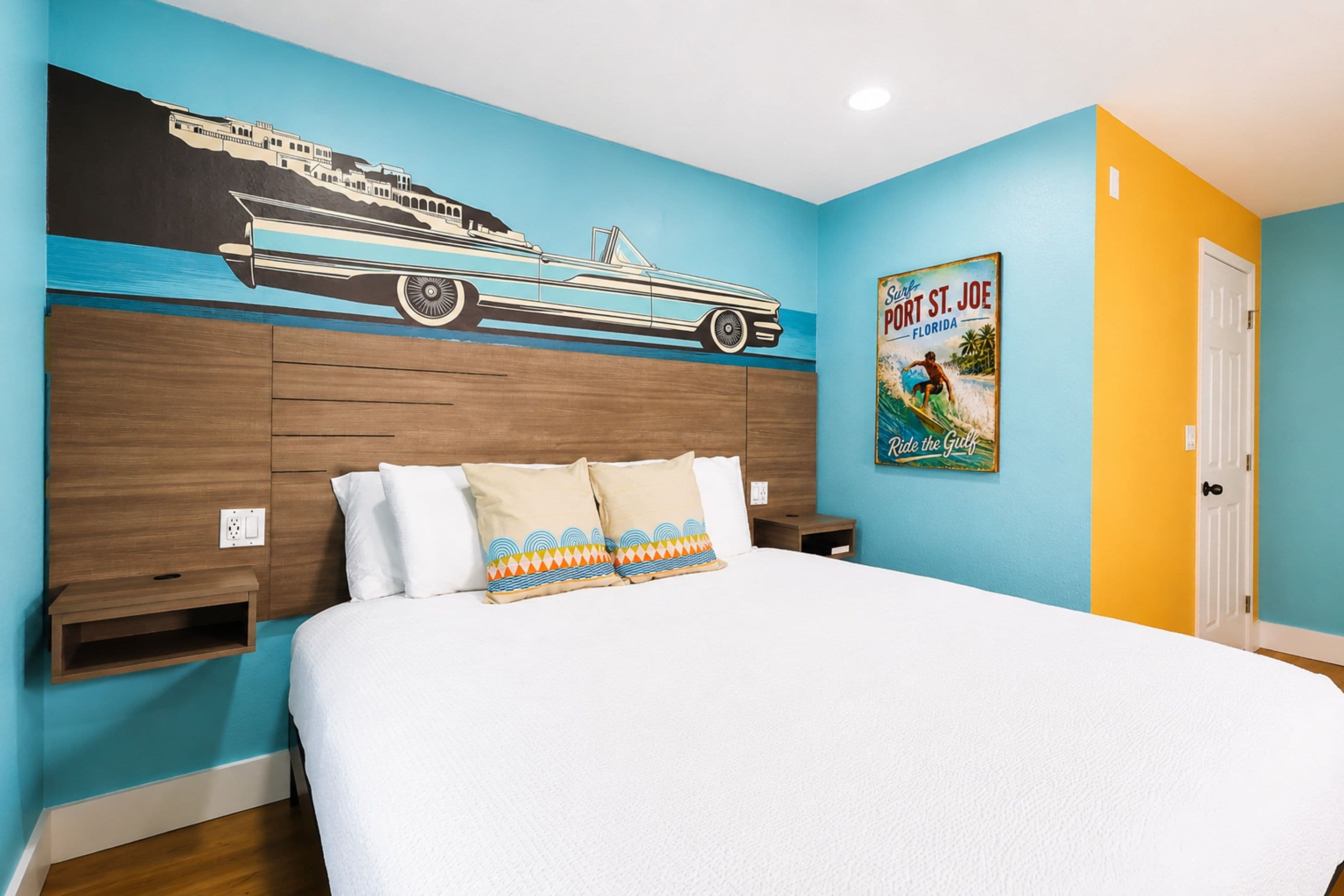 Quarto de hotel com uma cama grande, cabeceira de madeira, um pôster de um cruzeiro e um pôster de um surfista.