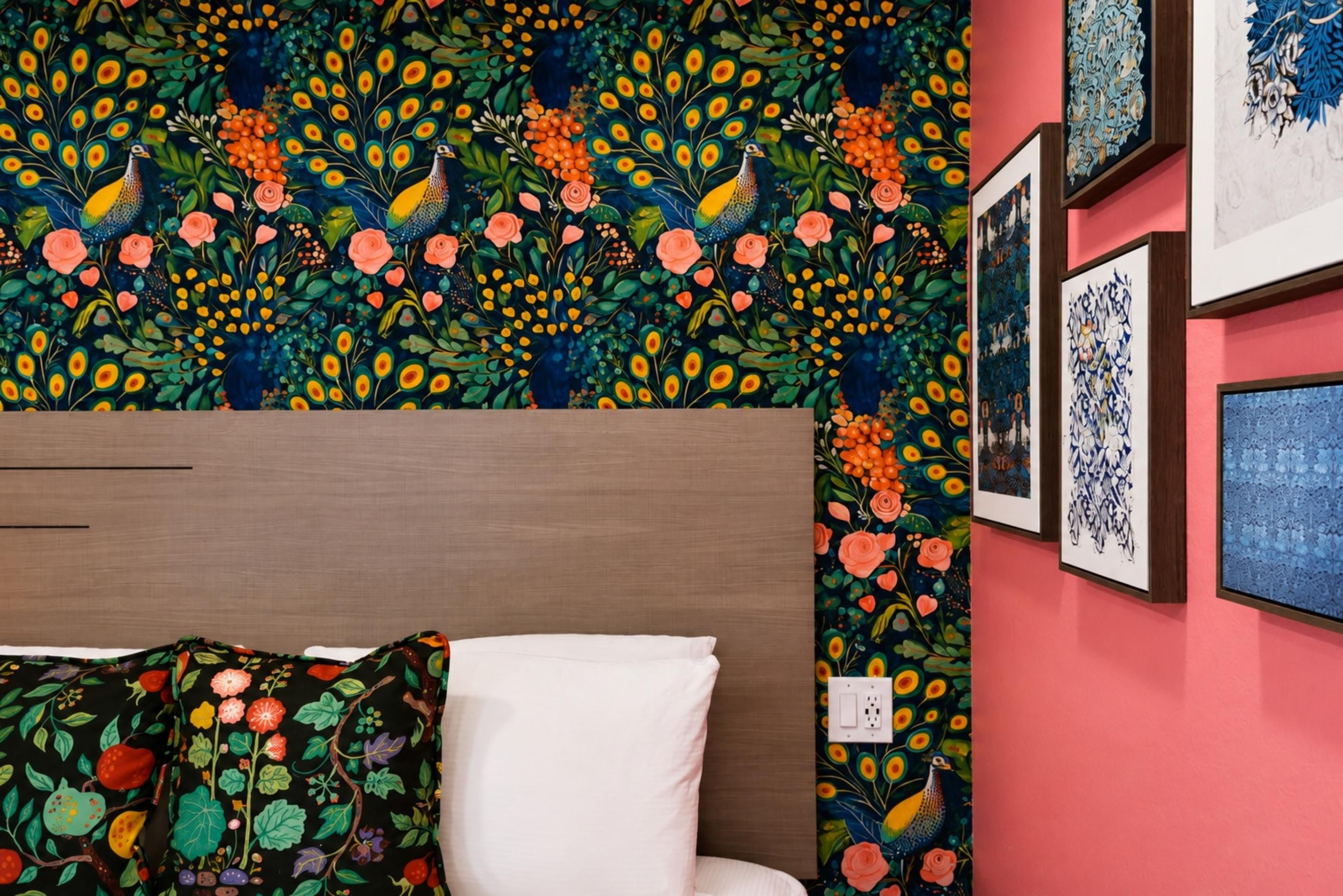 Quarto com papel de parede floral colorido, cabeceira de madeira, cama com travesseiro branco e diversas obras de arte emolduradas na parede.