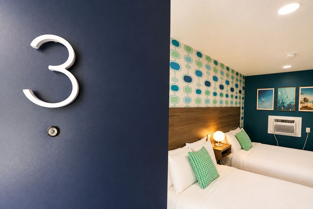 Quarto de hotel com o número '3' em destaque na parede, papel de parede estampado, cabeceira de madeira, duas camas com roupa de cama branca, almofadas verdes, mesa de cabeceira com abajur e ar condicionado fixado na parede.