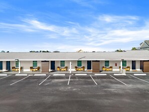Um motel com várias acomodações, cada uma com porta e janela. Há um estacionamento em frente ao motel.