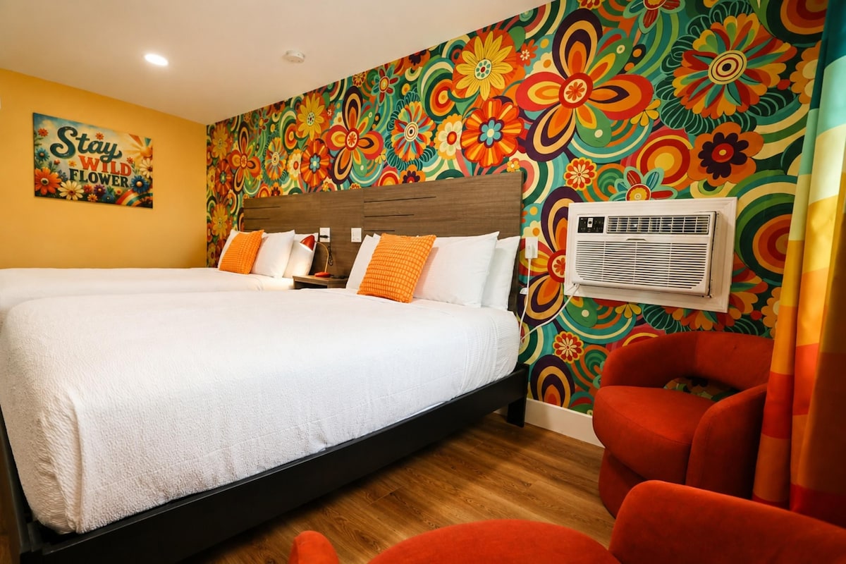 Quarto de hotel com uma cama grande, uma mesa de cabeceira, aparelho de ar condicionado e um vibrante papel de parede floral.