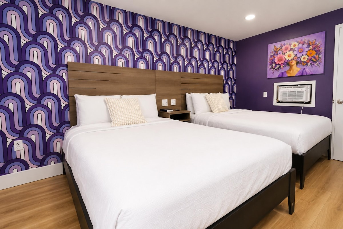 Quarto de hotel com duas camas, cabeceira de madeira, parede com padrão roxo e uma obra de arte floral.