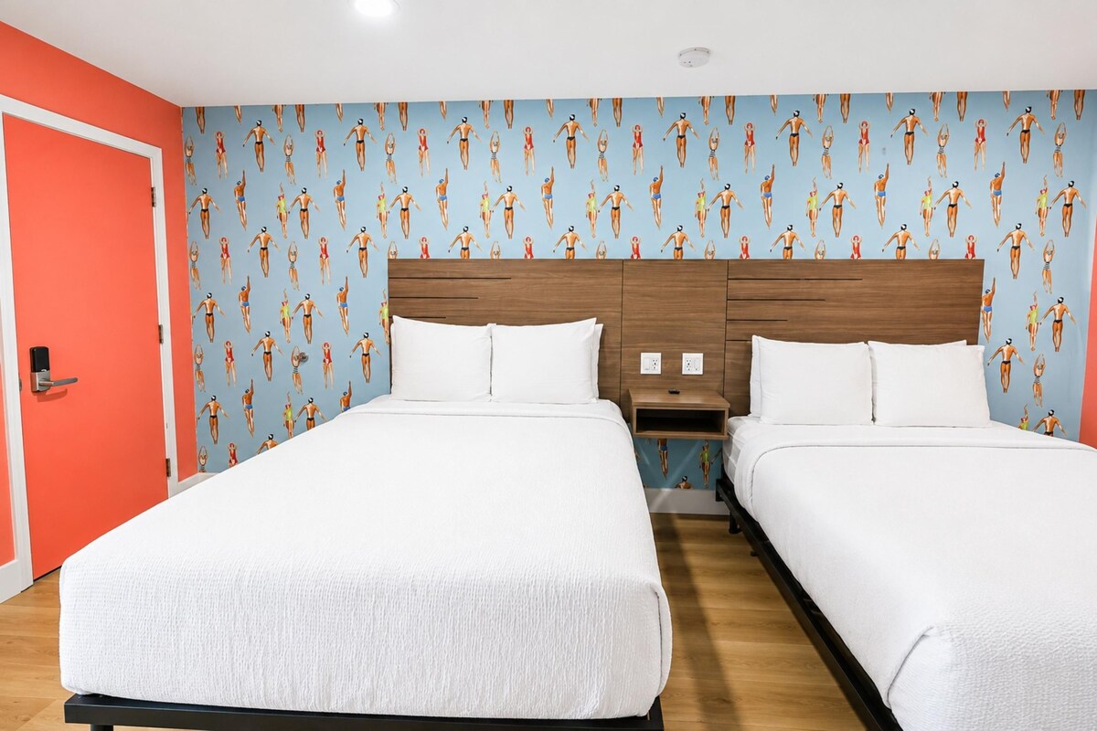 Quarto de hotel com duas camas, cabeceira de madeira, roupa de cama branca e parede decorada com um padrão de nadadores.