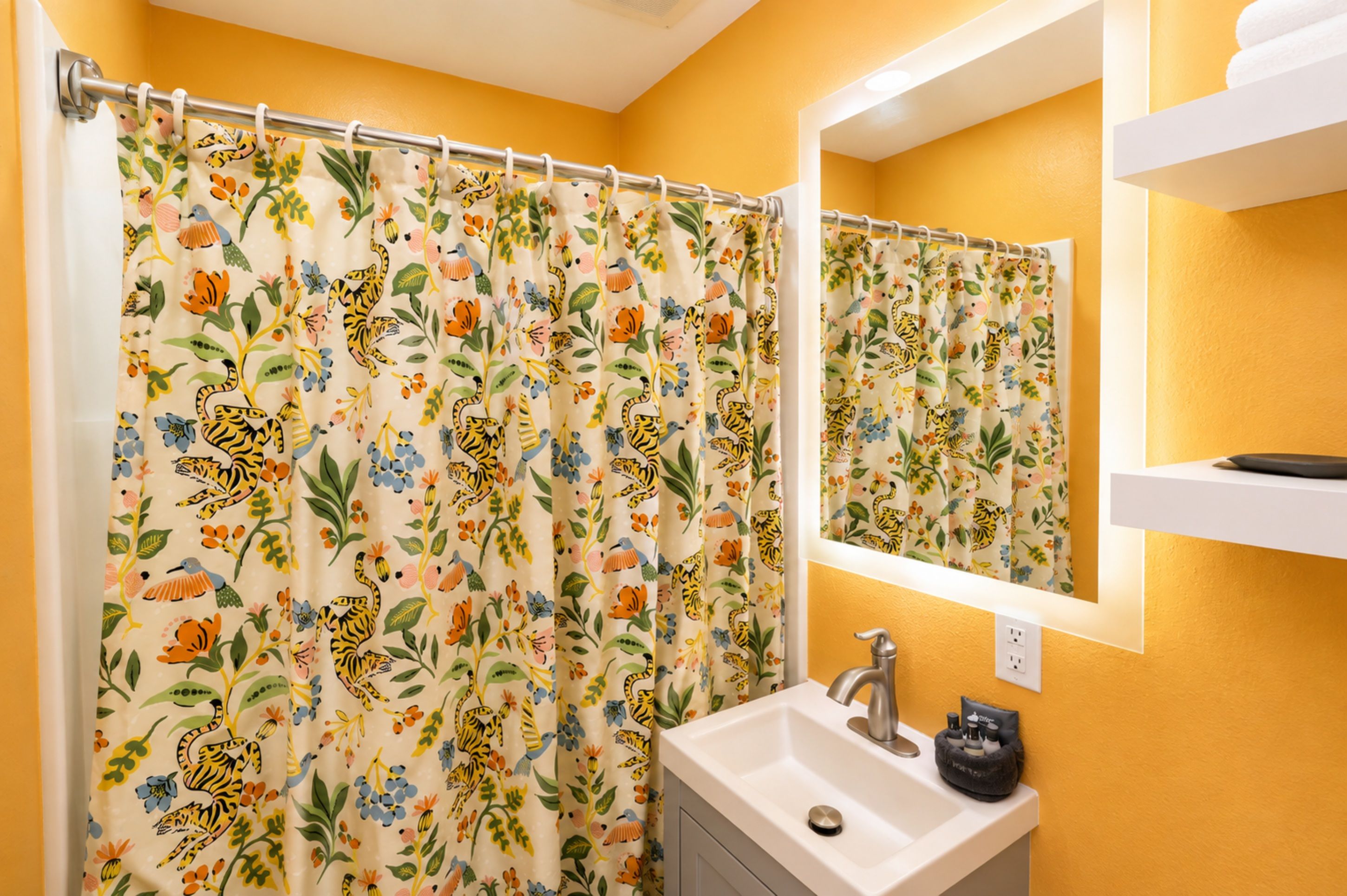 Casa de banho com cortina de chuveiro estampada com flores, pia com espelho em cima e prateleira com artigos de higiene pessoal.