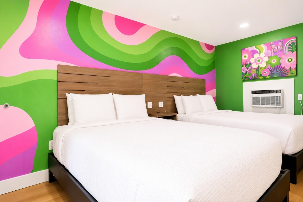 Quarto de hotel com duas camas, uma cabeceira de madeira, parede com um design abstrato colorido e uma pintura de flores.