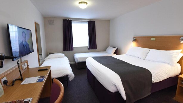 Quarto de hotel com uma cama, duas camas menores, uma escrivaninha com cadeira, televisão e uma janela com cortinas.