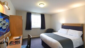 Quarto de hotel com cama, escrivaninha, cadeira, televisão e janela com cortinas.