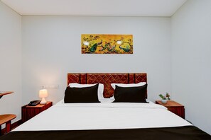 Quarto de hotel com cama, mesa de cabeceira com luminária, pintura na parede e pequena mesa com planta.