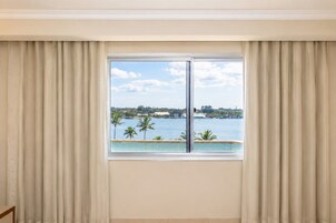 Quarto com uma grande janela, cortinas e vista para um corpo de água com palmeiras.