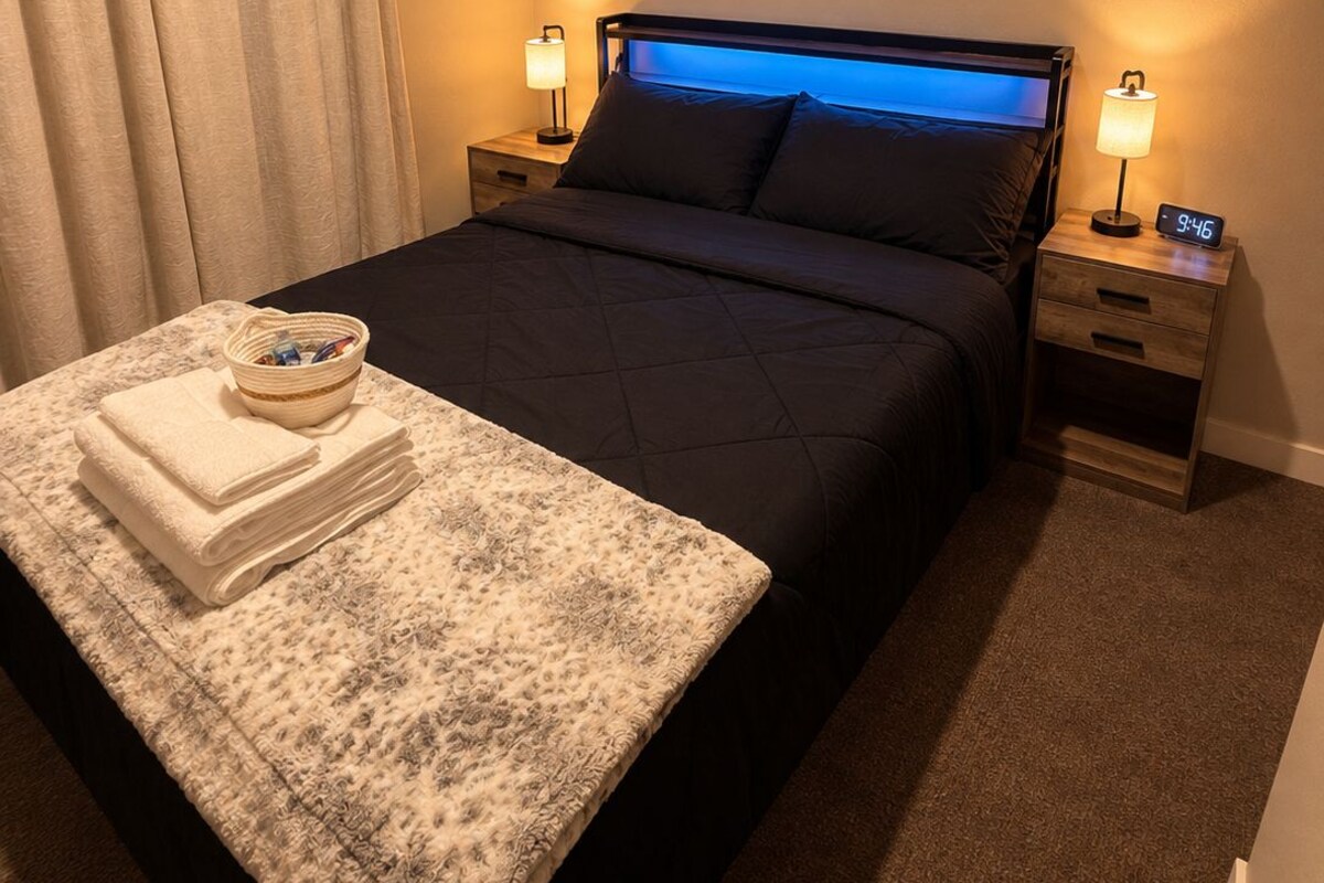 Quarto de hotel com cama, duas mesas de cabeceira, luminária, relógio, janela com vista e pilha de toalhas.