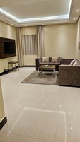 Uma sala de estar moderna com televisão de tela plana na parede, um sofá marrom com almofadas, uma mesa de centro de madeira e uma janela grande com cortinas.