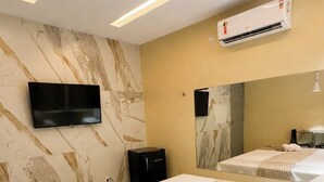 Quarto de hotel com televisão de tela plana na parede, cama com roupa de cama branca, tapete bege e aparelho de ar condicionado.