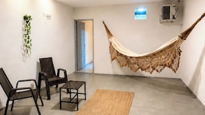 Quarto espaçoso com piso de concreto, uma rede, duas cadeiras, uma mesinha e uma planta.