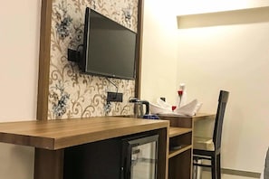 Quarto de hotel com escrivaninha de madeira, televisão na parede, pequena geladeira e área de refeições com cadeira e mesa.