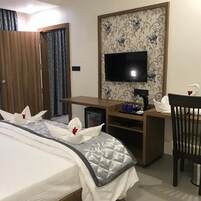 Quarto de hotel com cama, escrivaninha, cadeira, televisão e uma pequena mesa com objetos.