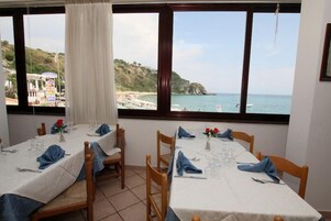 Uma área de refeições com mesas preparadas para dois, com vista para uma praia e um litoral rochoso.